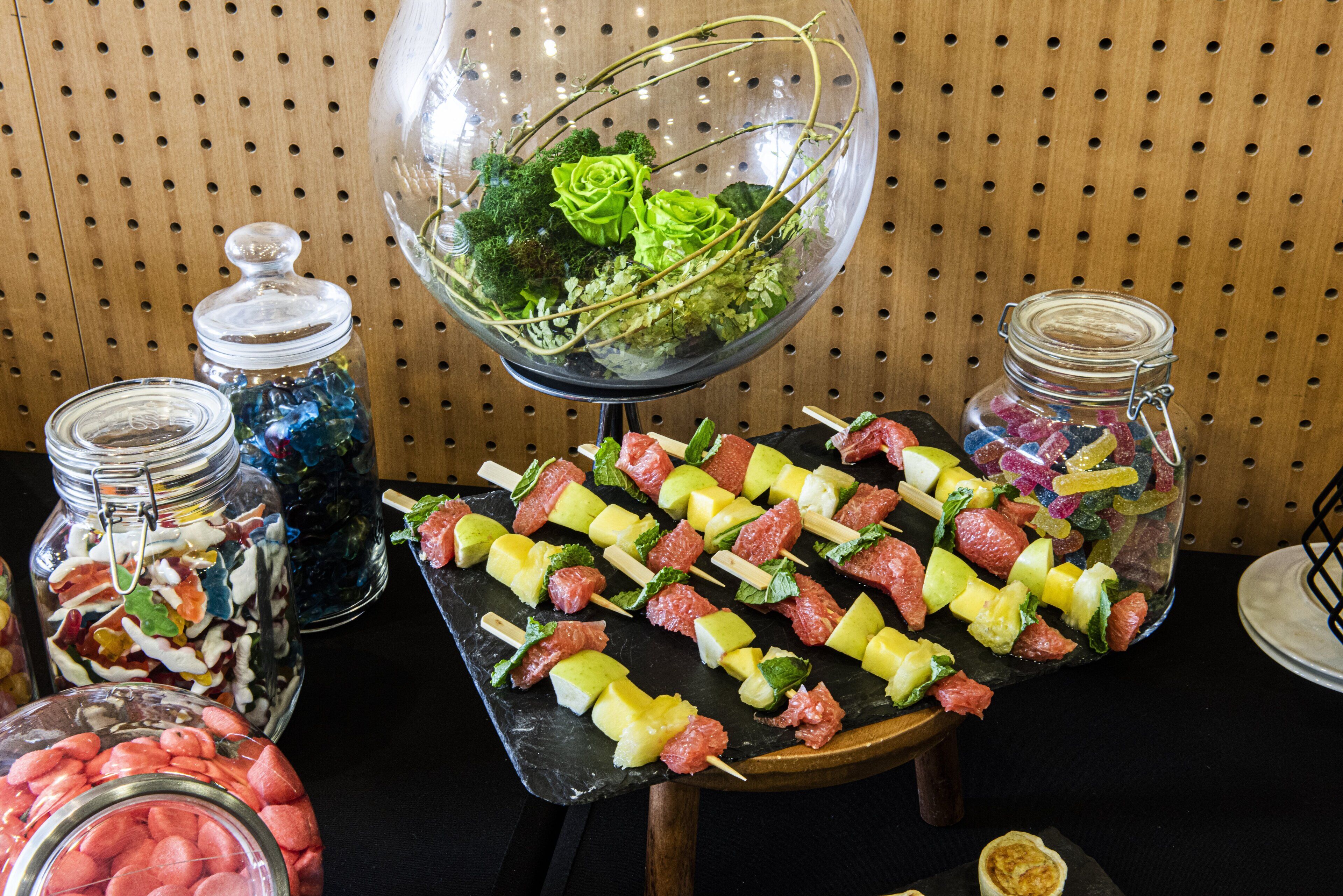 Exposição de comida com espetos de frutas e legumes, e potes com vários tipos de doces.