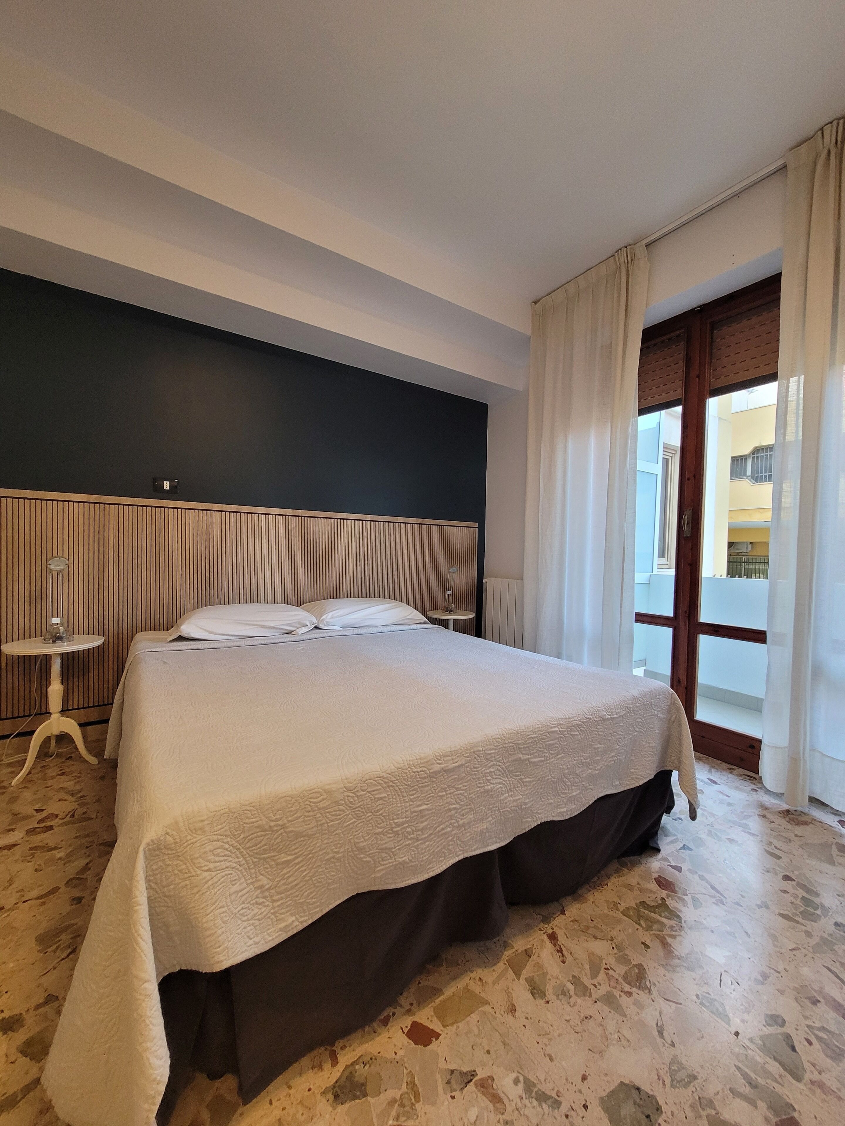 Quarto de hotel com uma cama, uma mesinha de cabeceira, uma luminária na parede e vista pela janela com cortinas.