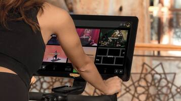 Mulher usando uma bicicleta ergométrica com tela digital exibindo programas de treino.