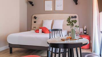 Quarto de hotel moderno com uma cama, uma mesa de jantar com cadeiras, um banquinho vermelho pequeno e uma planta em vaso.