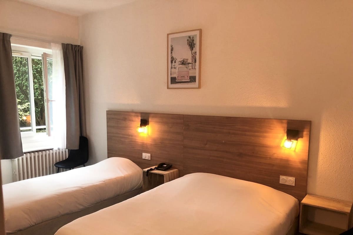 Quarto de hotel com duas camas de solteiro, cabeceira de madeira, luminárias de cabeceira, mesa de cabeceira pequena, uma cadeira, janela com cortinas e um quadro na parede.