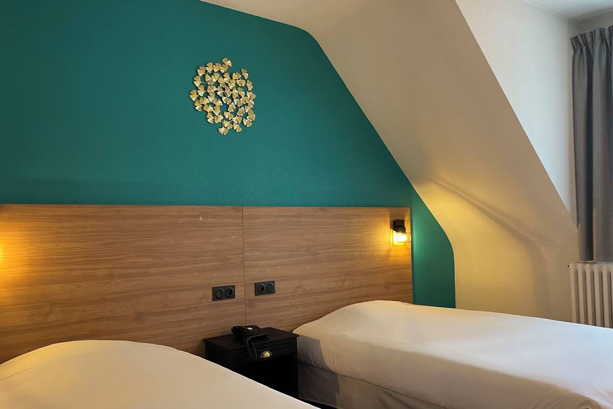 Quarto de hotel com duas camas, cabeceira de madeira, luminária fixada na parede e uma mesinha com telefone.