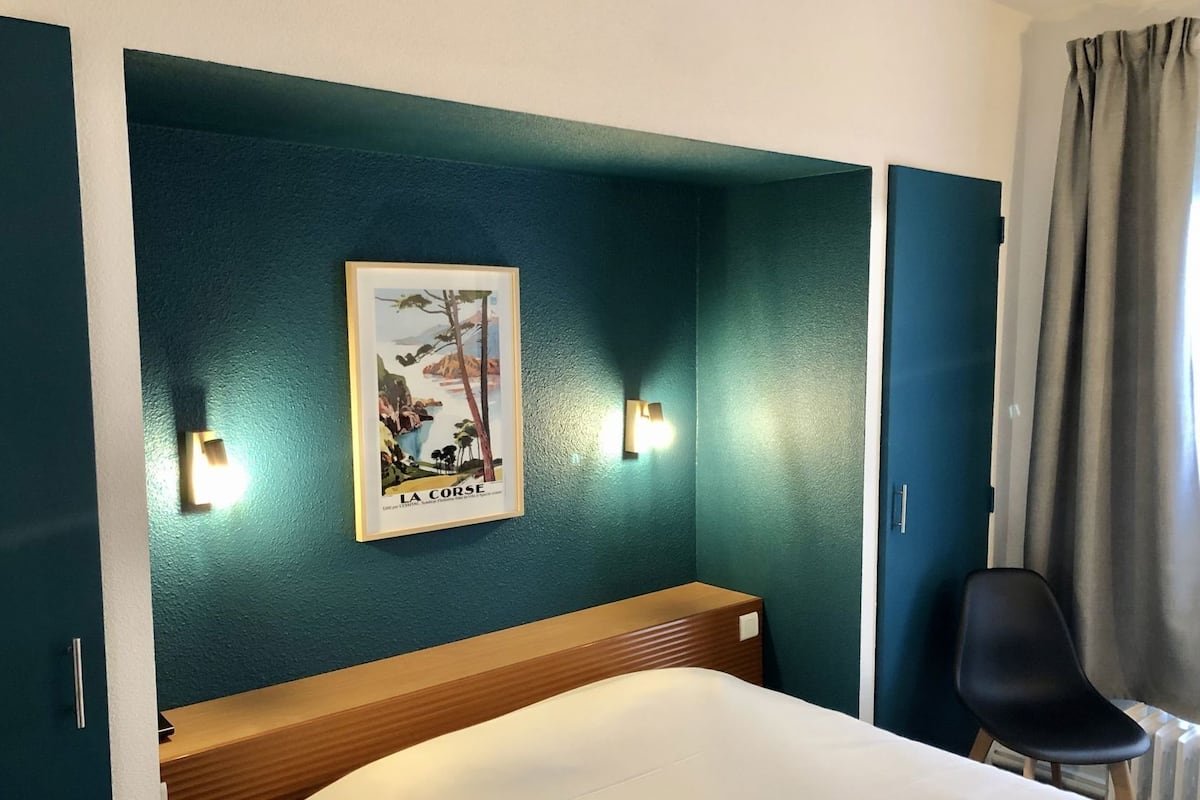 Quarto de hotel com cama, mesa de cabeceira, cadeira, luminária na parede e um quadro emoldurado.