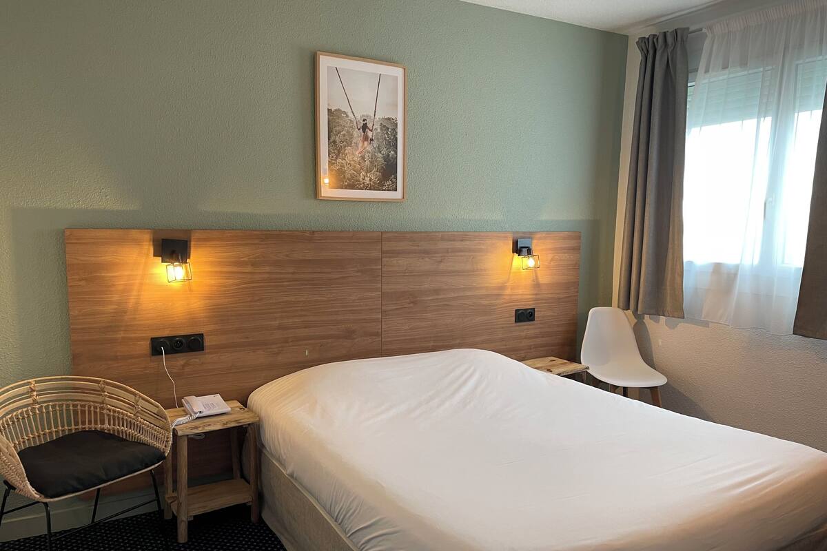 Quarto de hotel com uma cama grande, duas mesinhas de cabeceira, uma cadeira, um quadro na parede e uma janela com cortinas.