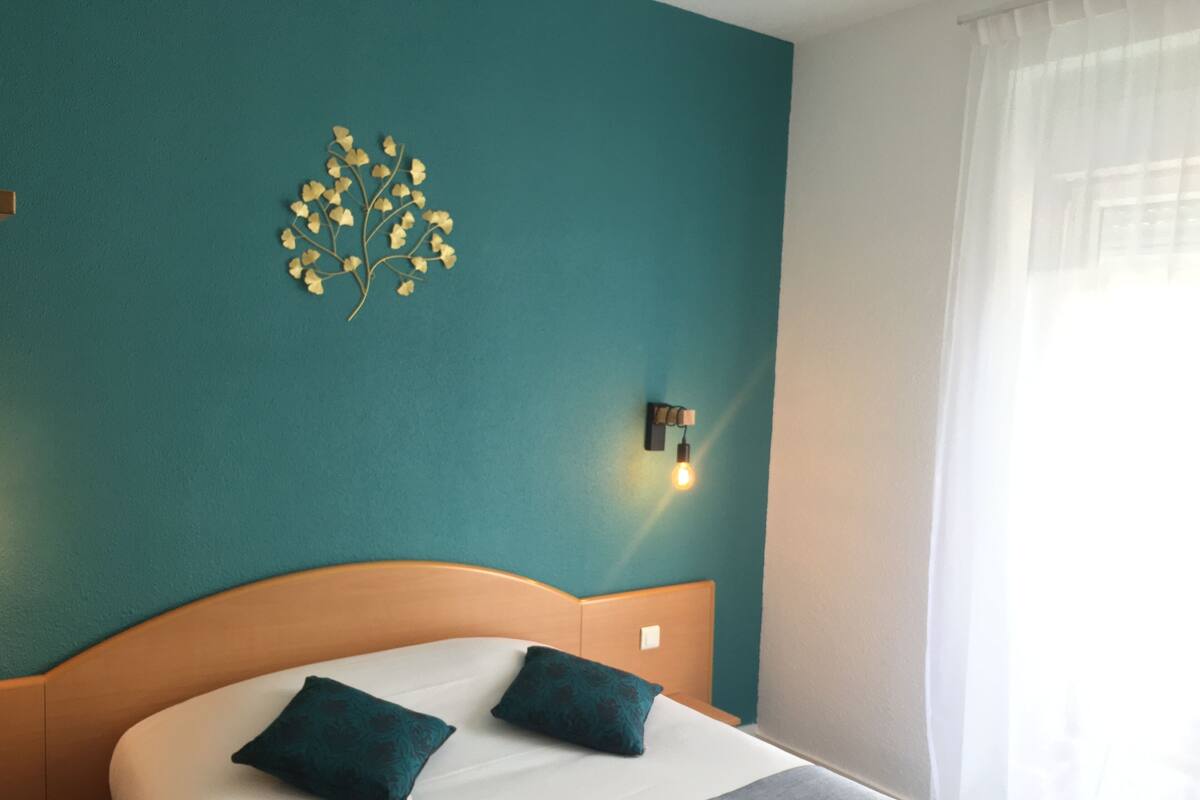 Uma cama bem arrumada com duas almofadas, uma luminária fixada na parede e um objeto decorativo na parede.
