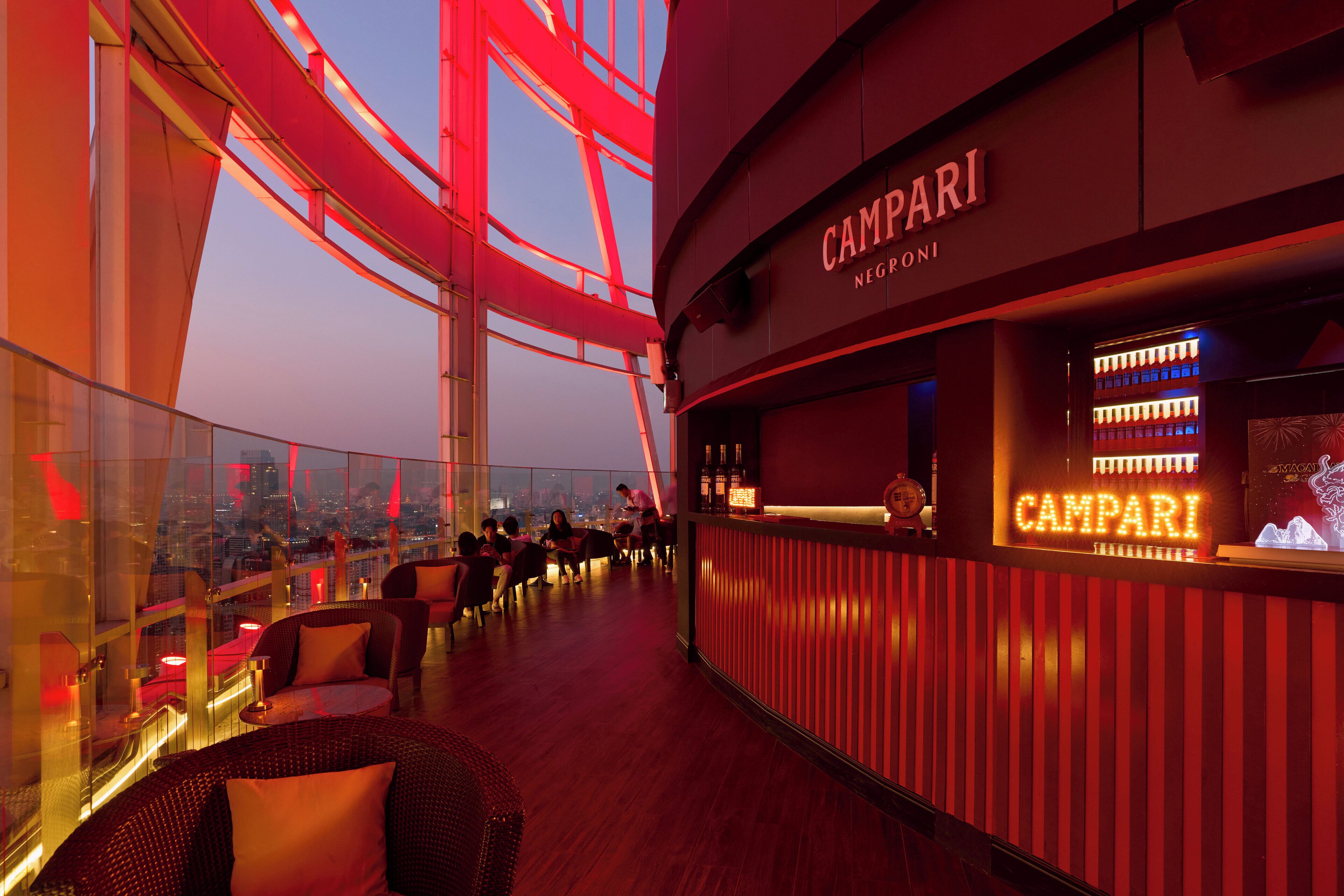 Um bar no terraço com vista para a cidade, com interior iluminado em vermelho e um letreiro 'Campari'.