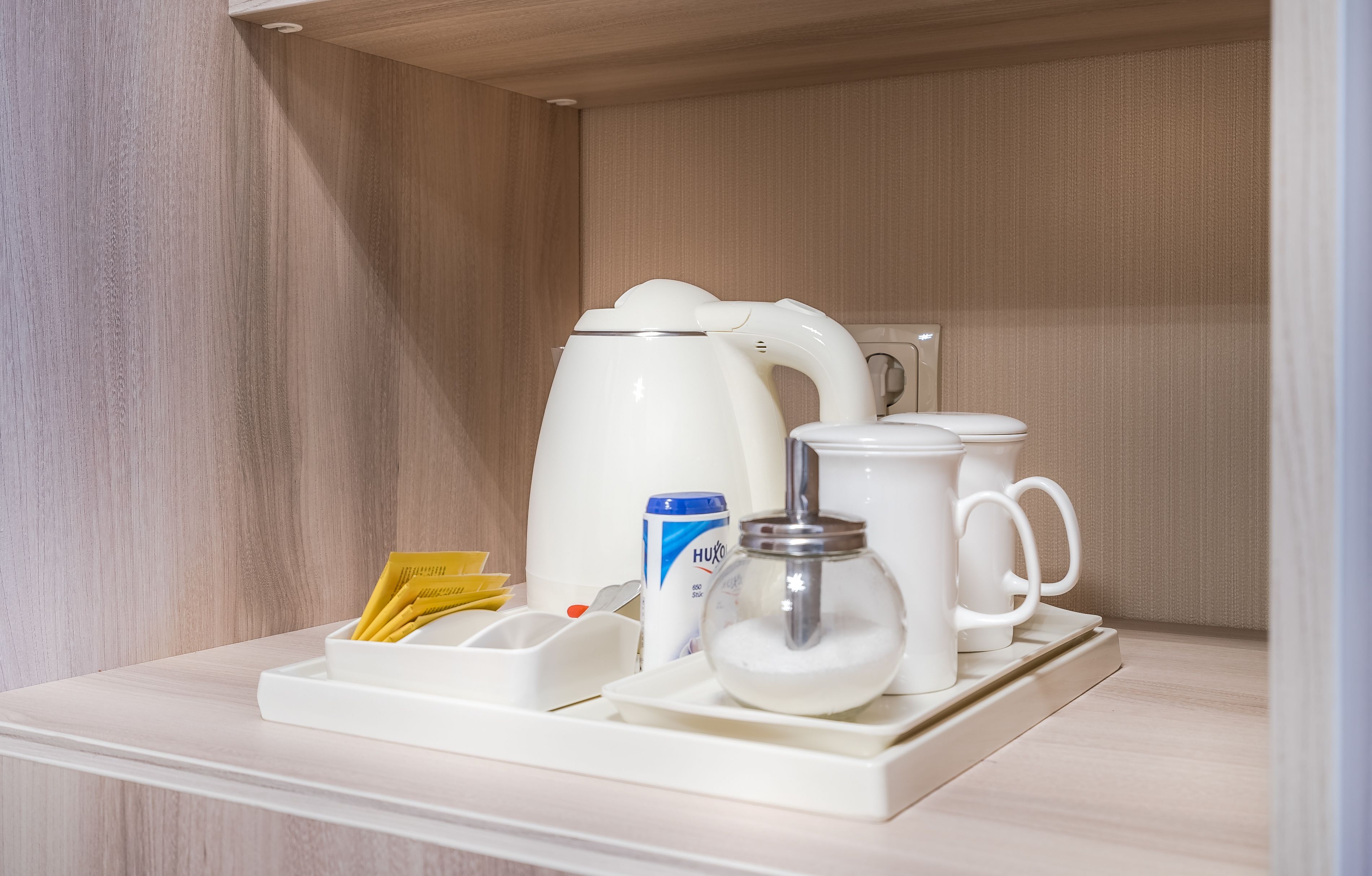 Uma área de cozinha com cafeteira, chaleira, caneca, pote de vidro e uma caixa de guardanapos amarelos em uma bancada branca.