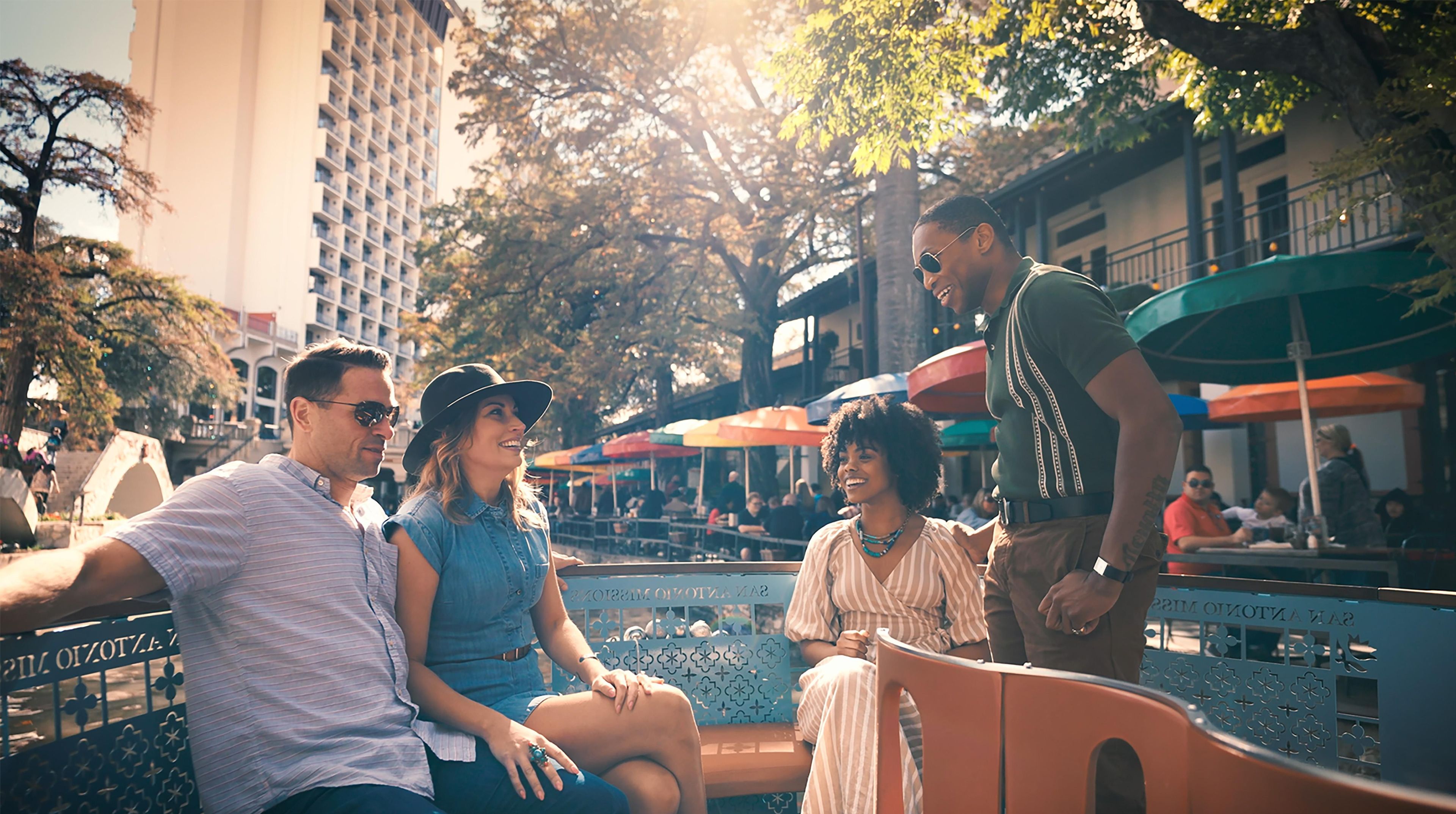 Um grupo de pessoas sentadas num banco em ambiente urbano ao ar livre, com edifícios e árvores ao fundo.