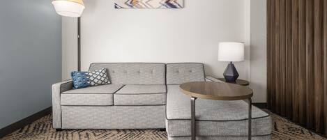 Quarto moderno com sofá cinza, mesa lateral redonda de madeira, luminária de chão e tapete estampado.