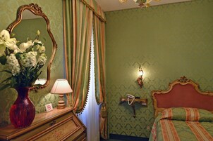 Um quarto com cama, cômoda, espelho, vaso com flores, abajur, cortinas e telefone fixado na parede.