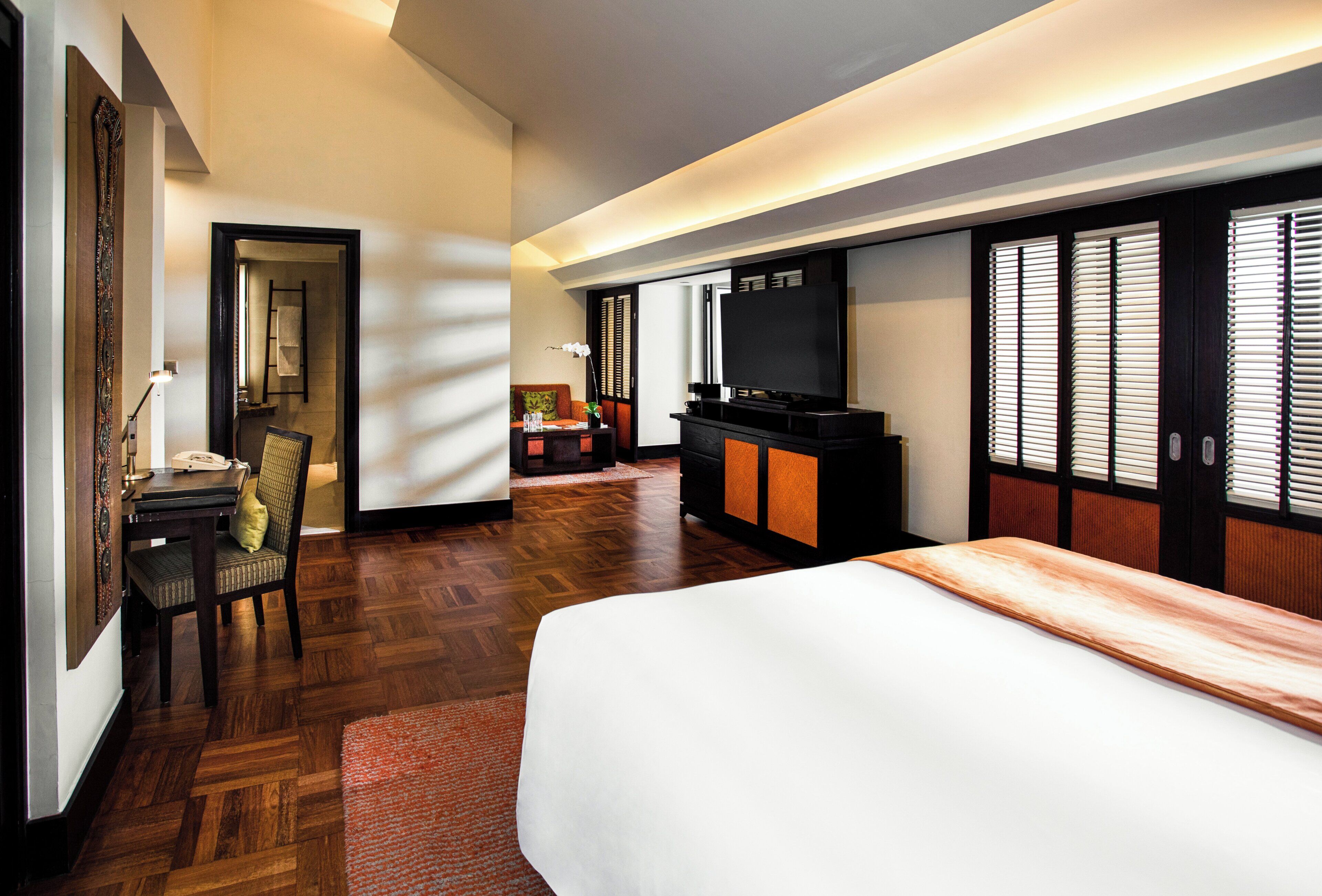 Quarto de hotel com cama, escrivaninha com cadeira, televisão e uma janela grande com persianas.