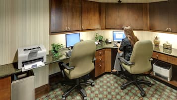 Escritório de hotel com duas mesas, computadores, cadeiras, uma impressora e plantas decorativas.