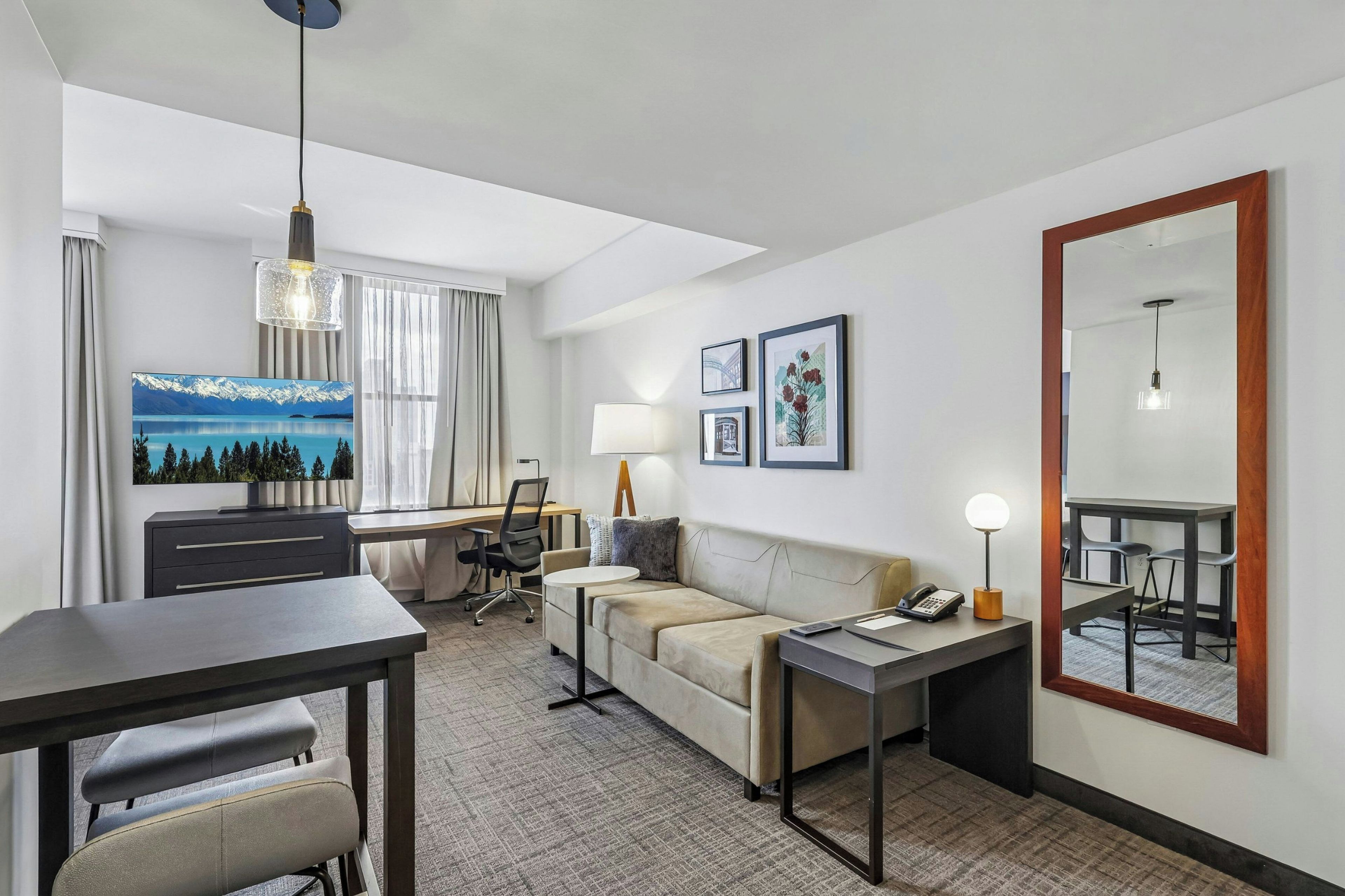 Quarto de hotel com escrivaninha, sofá, mesa de jantar, cadeira, luminária e um espelho grande.
