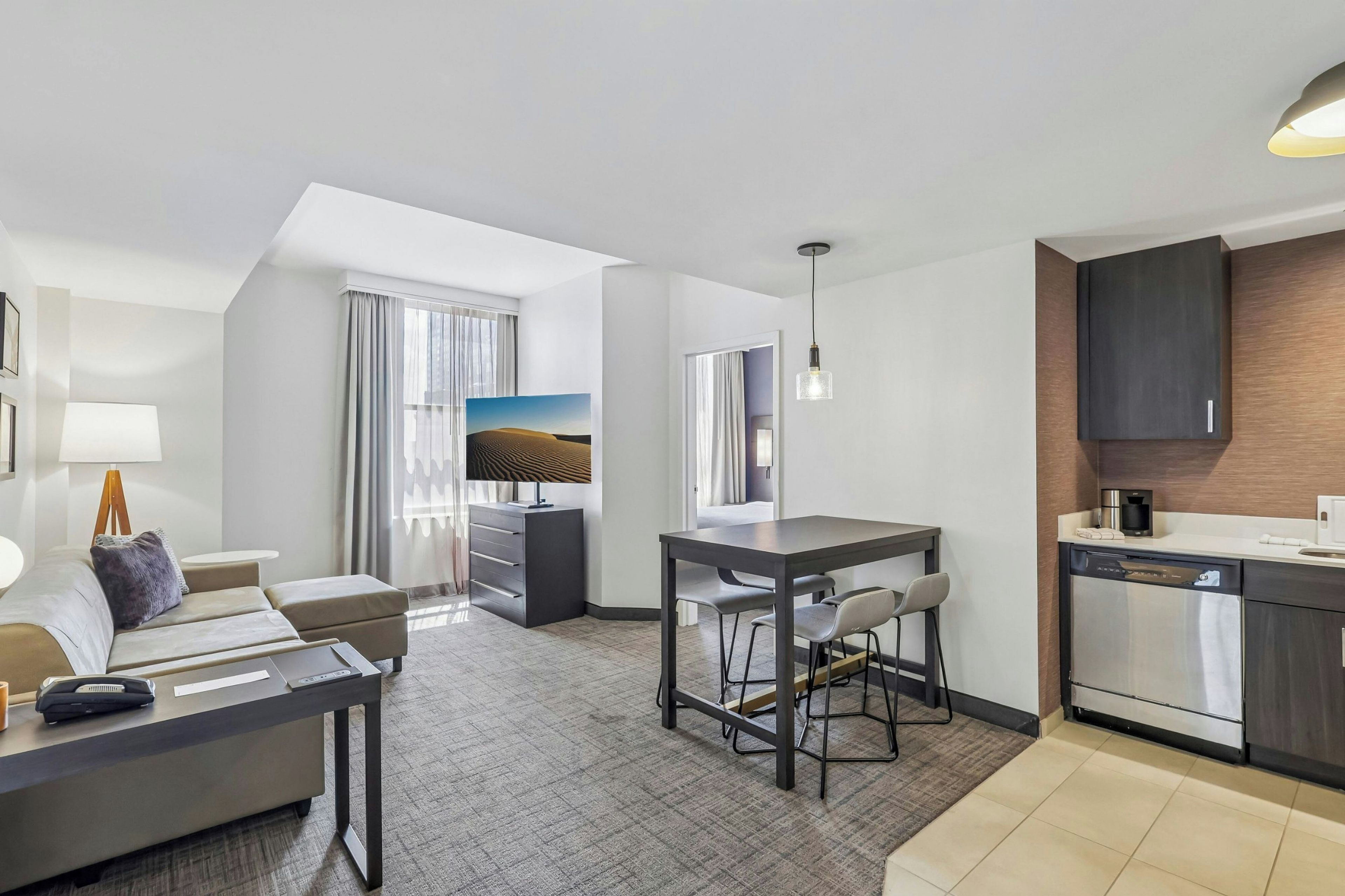 Quarto de hotel moderno com sofá, mesa de jantar com cadeiras, cozinha e uma janela grande com cortinas.