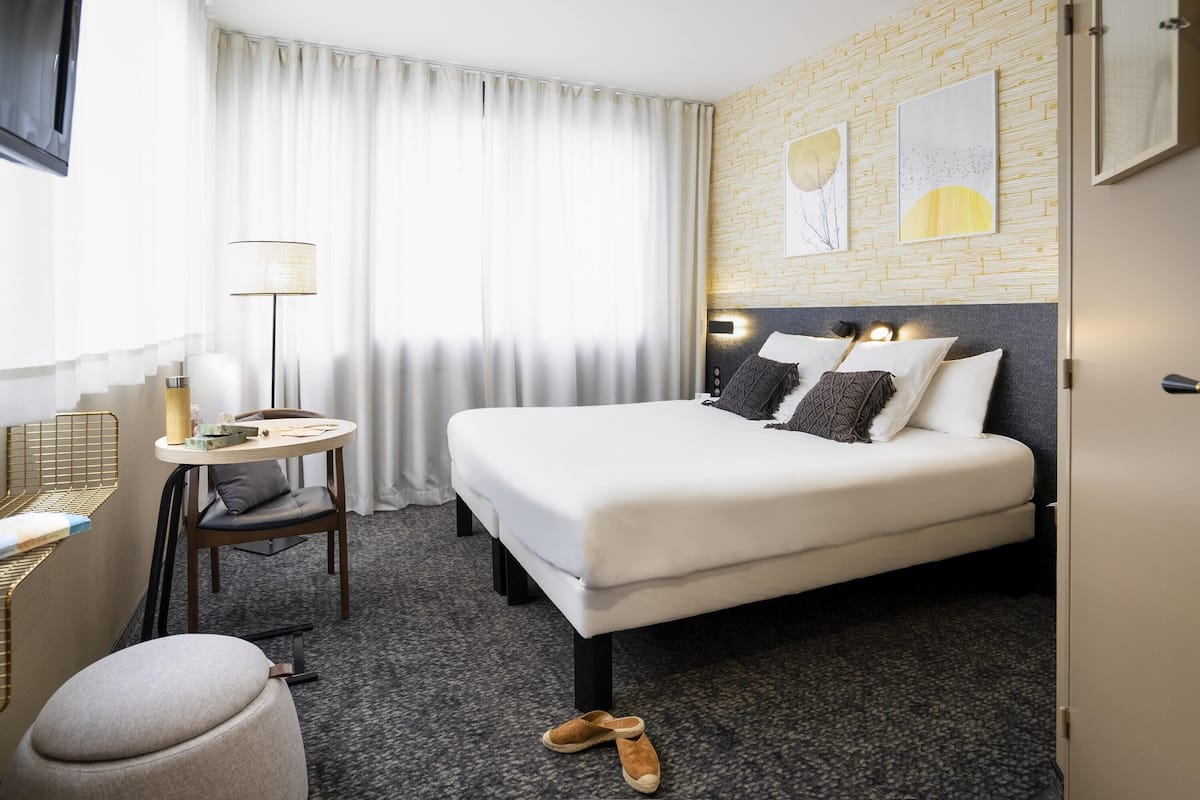 Quarto de hotel com cama, escrivaninha, cadeira, abajur e parede decorada com obras de arte.