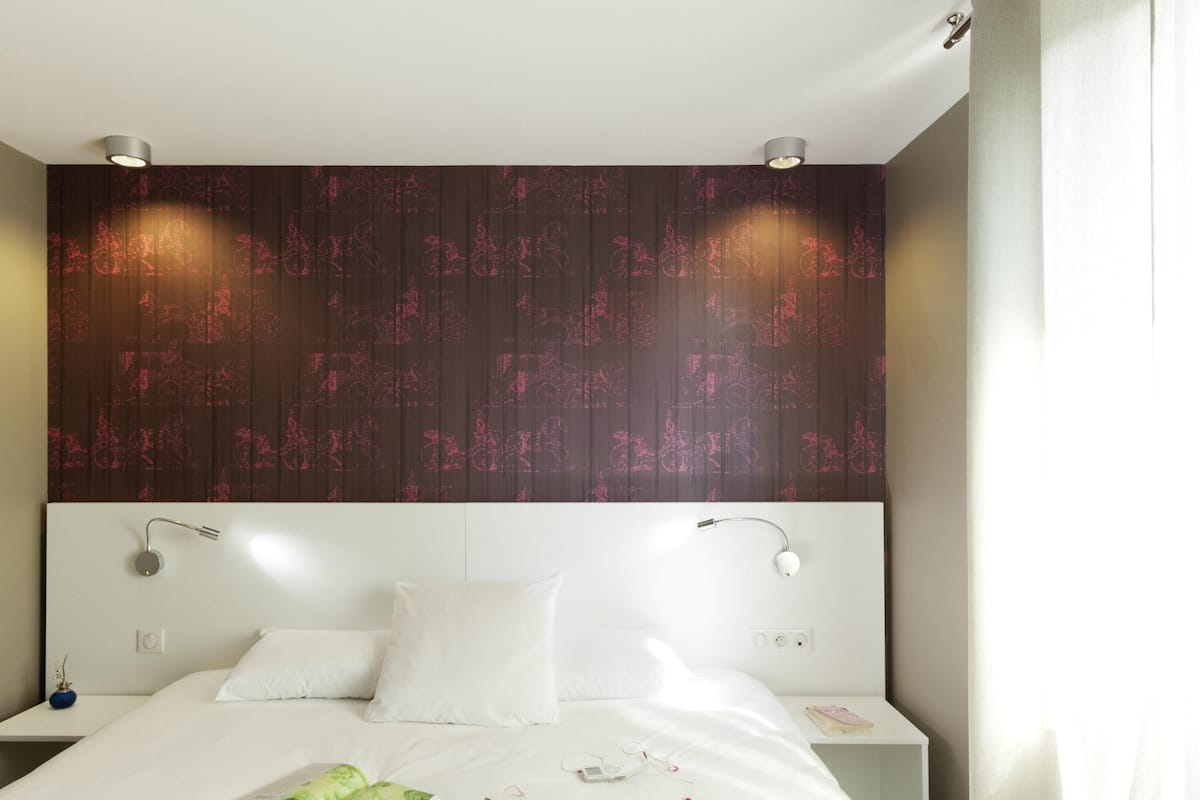 Quarto de hotel moderno com uma cama grande, roupa de cama branca, cabeceira com design estampado, mesas de cabeceira e luminária fixada na parede.