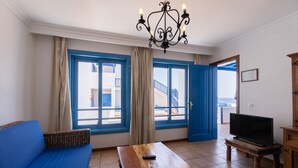 Uma sala de estar com um sofá azul, uma mesa de centro de madeira, uma televisão sobre um móvel de madeira e janelas amplas com molduras azuis.
