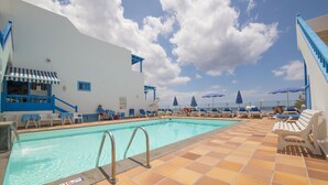 Uma área de piscina com cadeiras e guarda-sóis azuis e brancos, e uma escadaria que dá para um terraço superior.