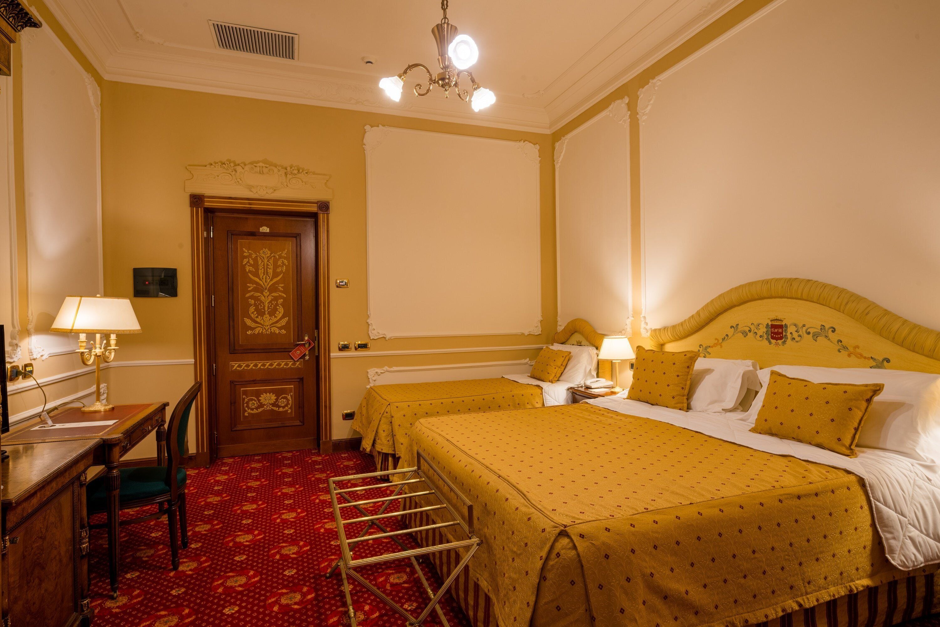Quarto de hotel com duas camas, uma escrivaninha, uma cadeira, um tapete com padrão vermelho e um lustre.