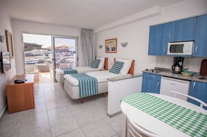 Quarto de hotel com duas camas, cozinha compacta, área de refeições e varanda com vista para a cidade.