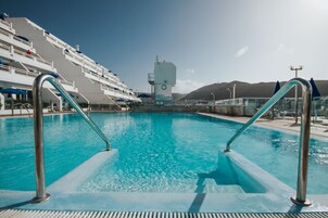 Piscina de navio de cruzeiro com grades de metal, água azul cristalina e um deck branco com degraus que levam à piscina.