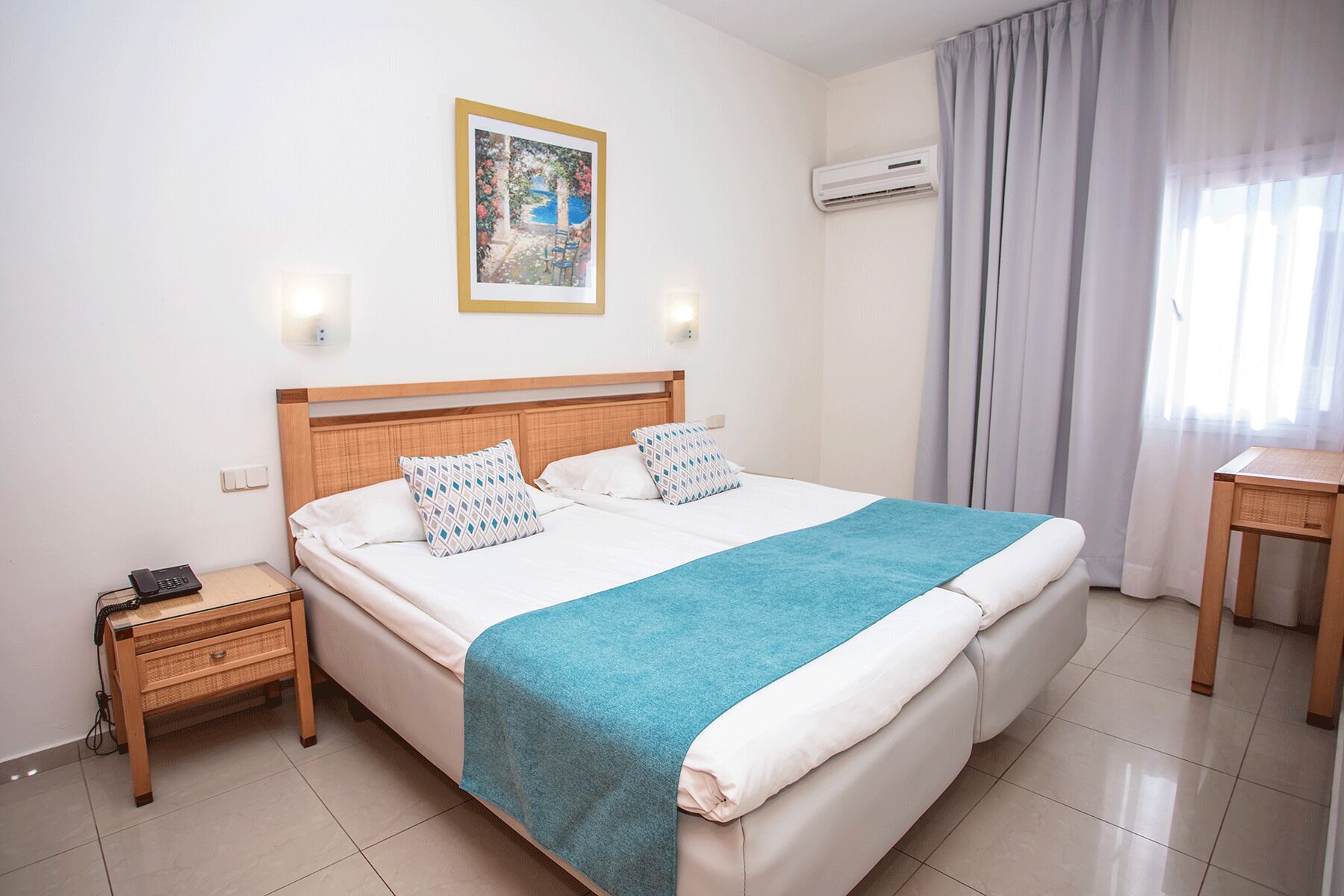 Cama bem arrumada com manta azul, mesa de cabeceira com telefone e um quadro na parede.