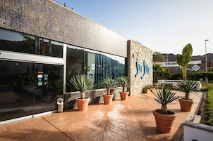 Entrada de hotel com pátio ladrilhado, plantas em vasos e uma placa com o nome do hotel.