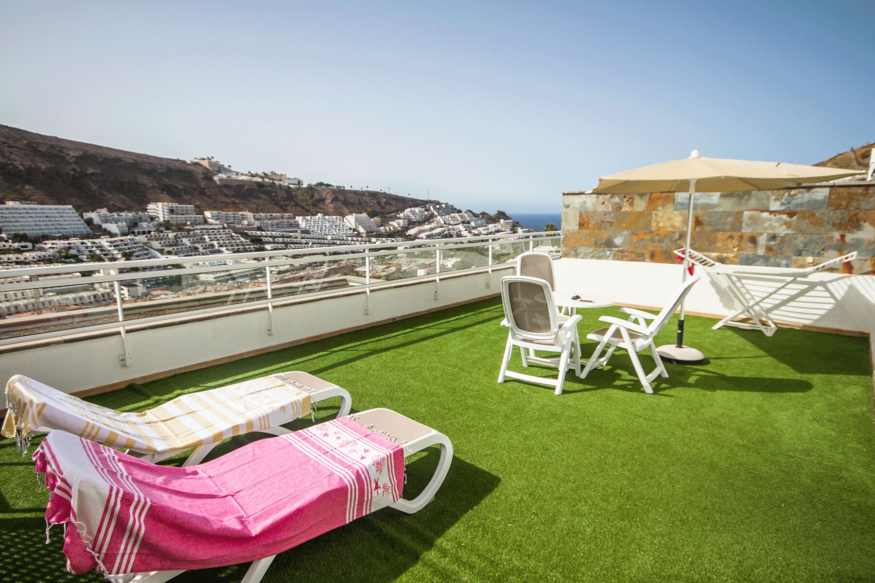 Um terraço no telhado com duas cadeiras de descanso, uma mesa e um guarda-sol. O terraço oferece uma vista desobstruída de uma cidade costeira e do mar.