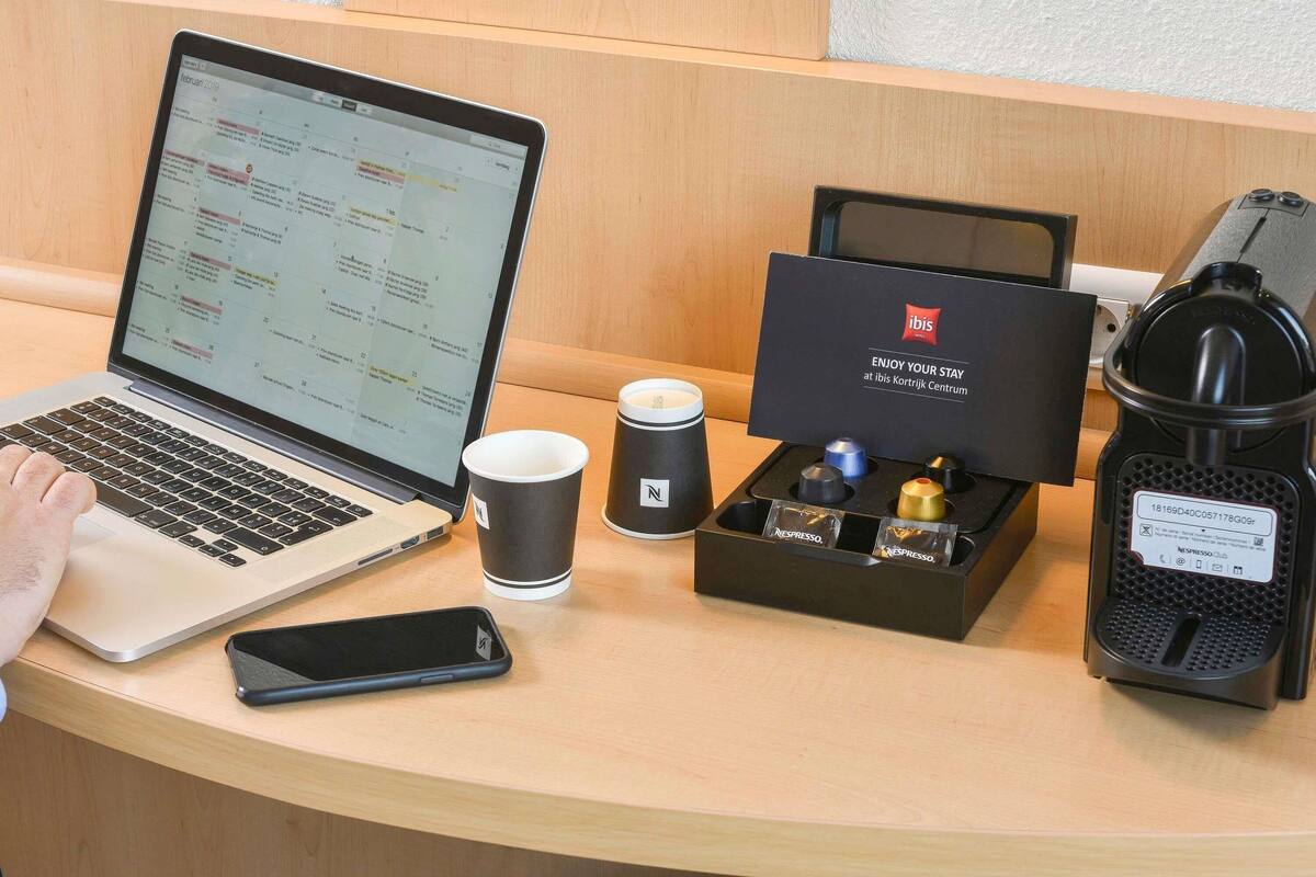 Um laptop, um smartphone, xícaras de café, uma máquina de café Nespresso e uma placa com o nome Ibis Kortrijk Centrum.