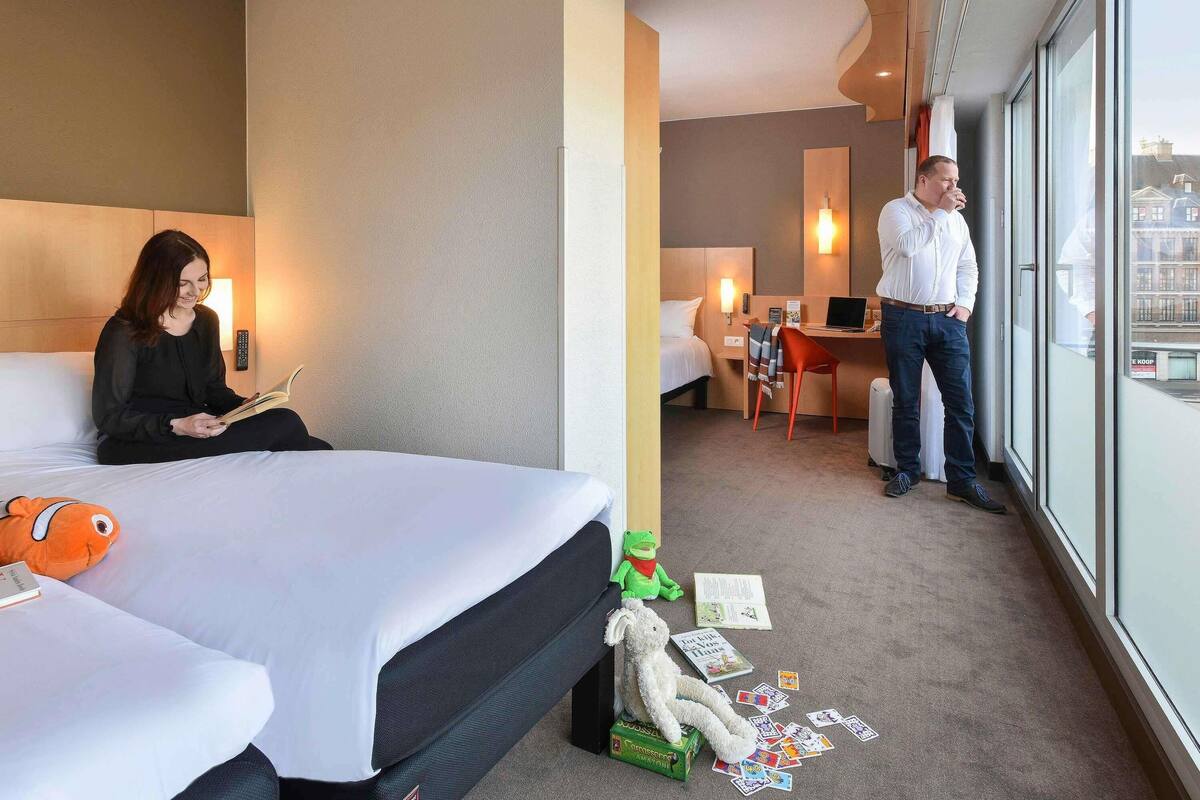 Quarto de hotel com cama, escrivaninha e vista da cidade por grandes janelas.