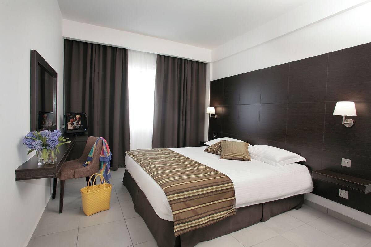 Quarto de hotel com cama, escrivaninha com cadeira, luminária e uma grande janela com cortinas.
