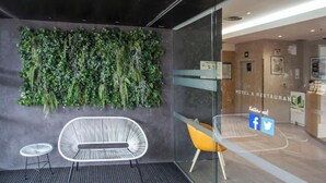 Um hall de hotel moderno com uma parede de jardim vertical, uma área de estar diferenciada e um painel de vidro com ícones de redes sociais.