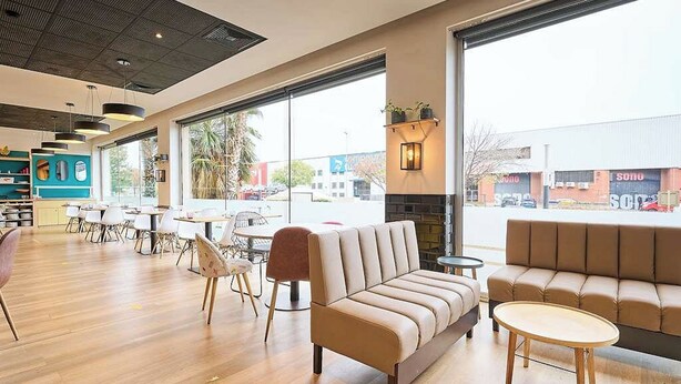 Um café amplo com interior moderno, grandes janelas e diversas opções de assento, incluindo sofás e mesas.