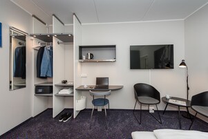 Quarto de hotel compacto com escrivaninha, cadeira e TV na parede.