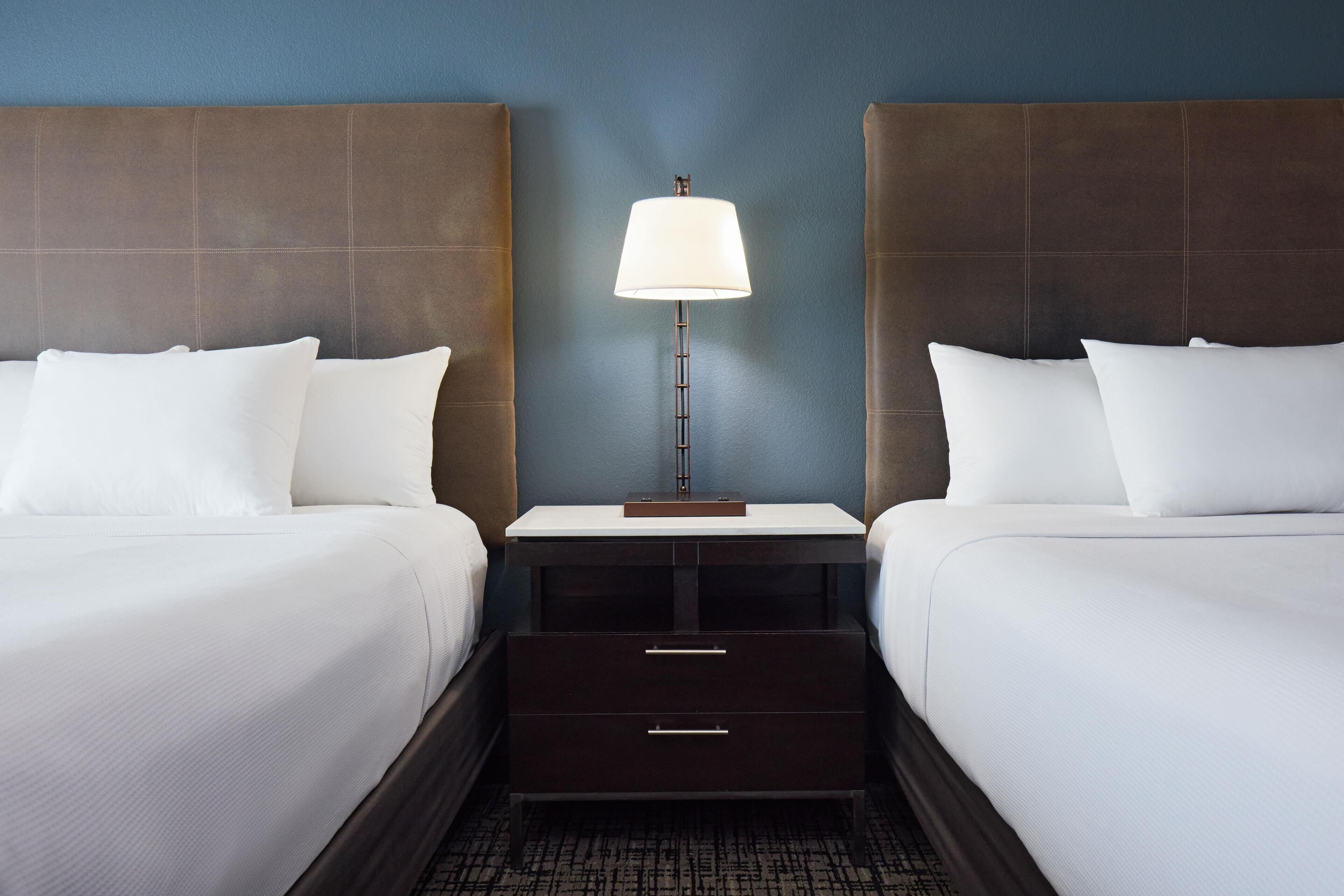 Quarto de hotel com duas camas, uma mesa de cabeceira com abajur e cabeceira de madeira escura.