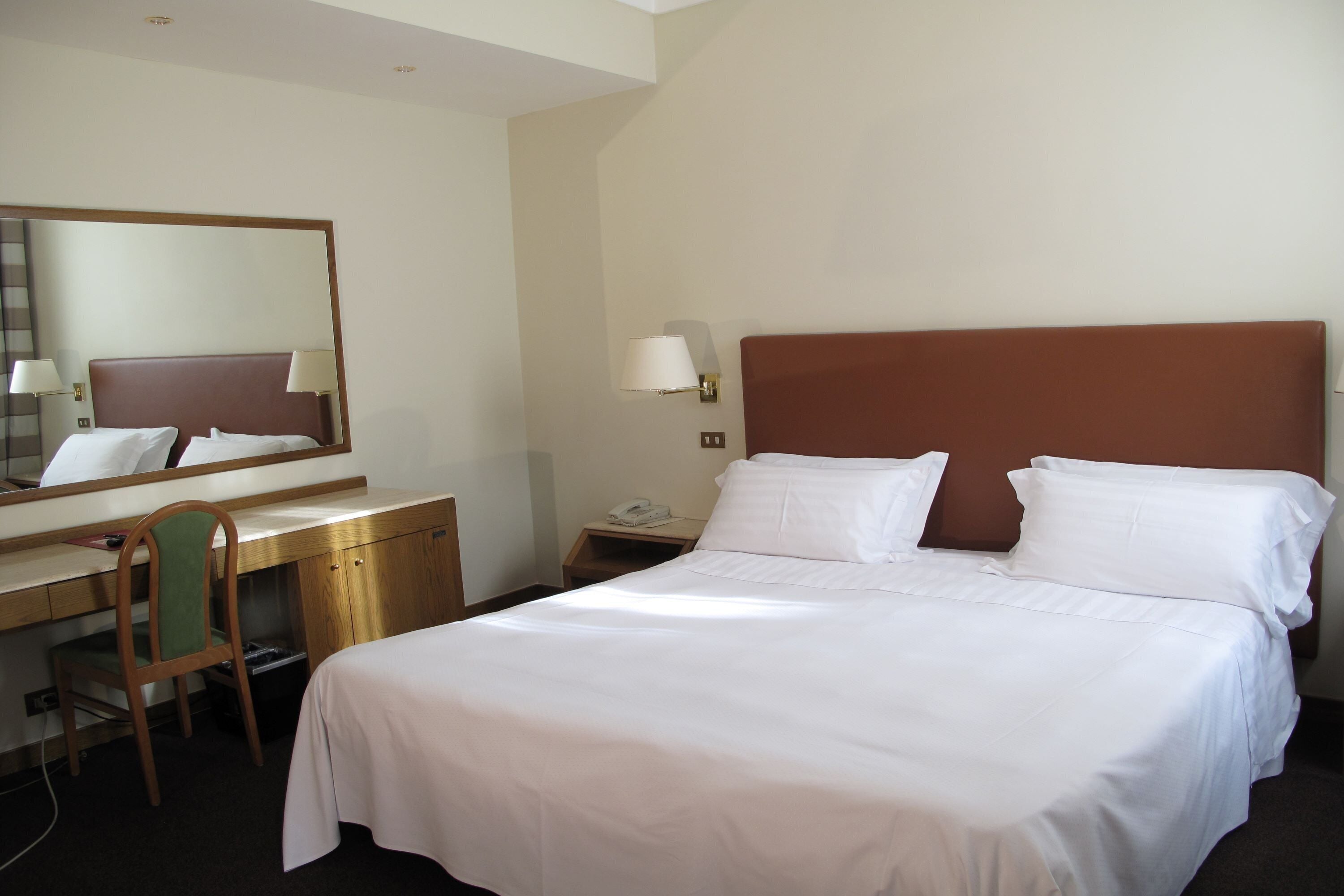 Quarto de hotel com uma cama de solteiro, uma mesa com cadeira, telefone e espelho.