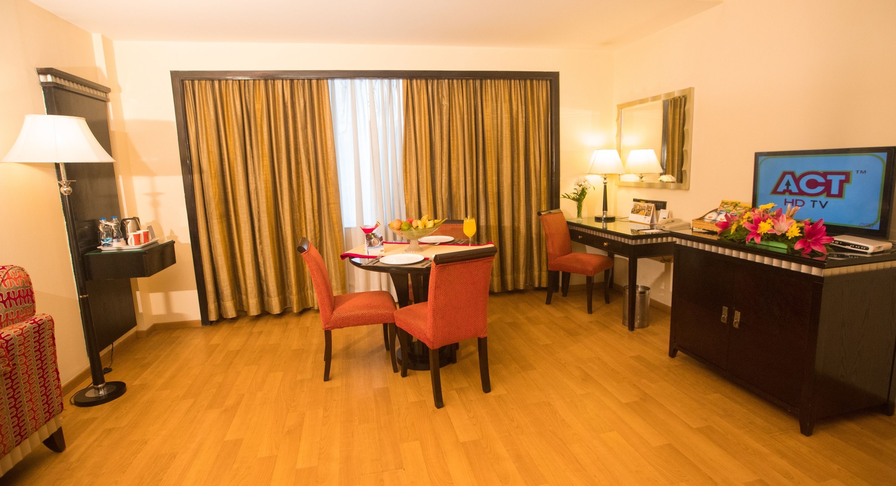 Quarto de hotel com uma mesa de jantar, duas cadeiras, televisão, escrivaninha, luminária e cama com cobertura estampada.