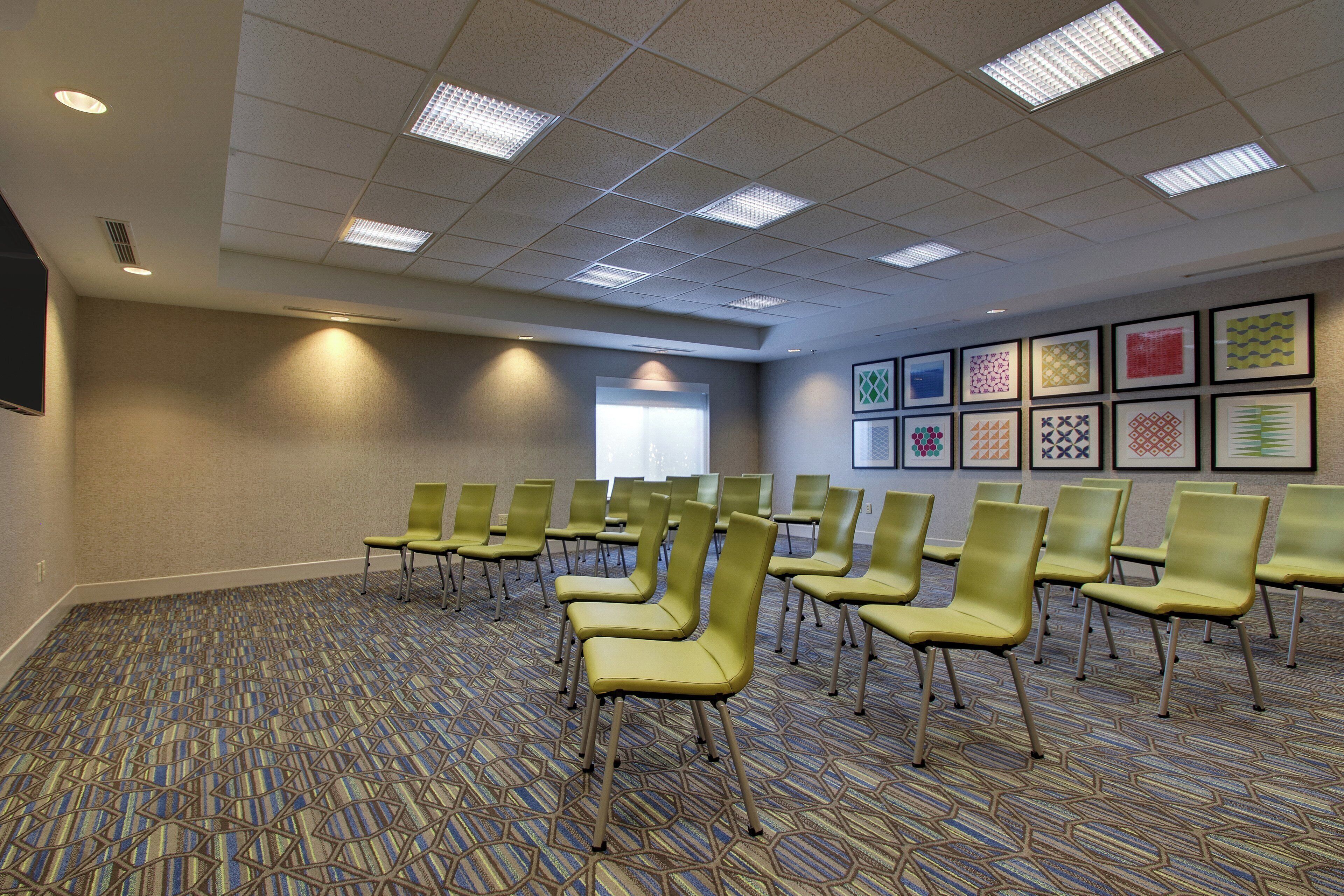 Sala de conferências com fileiras de cadeiras, carpete estampado e luminárias no teto.
