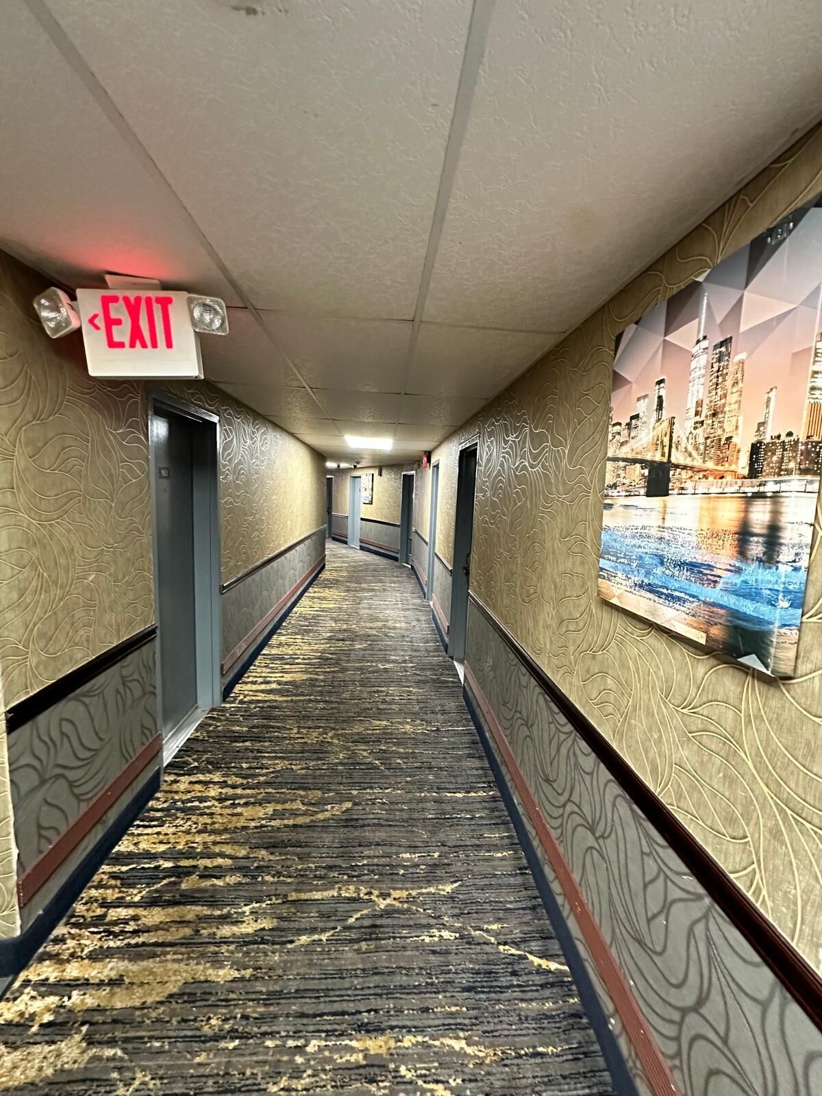 Corredor de hotel com paredes estampadas, placa de saída e um mural com vista para a cidade.
