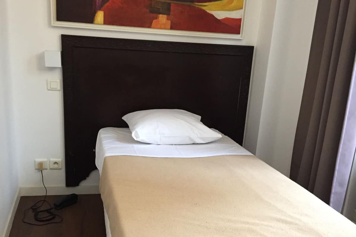 Uma cama de solteiro com cabeceira branca e cobertor bege, uma tomada e um cabo no chão.