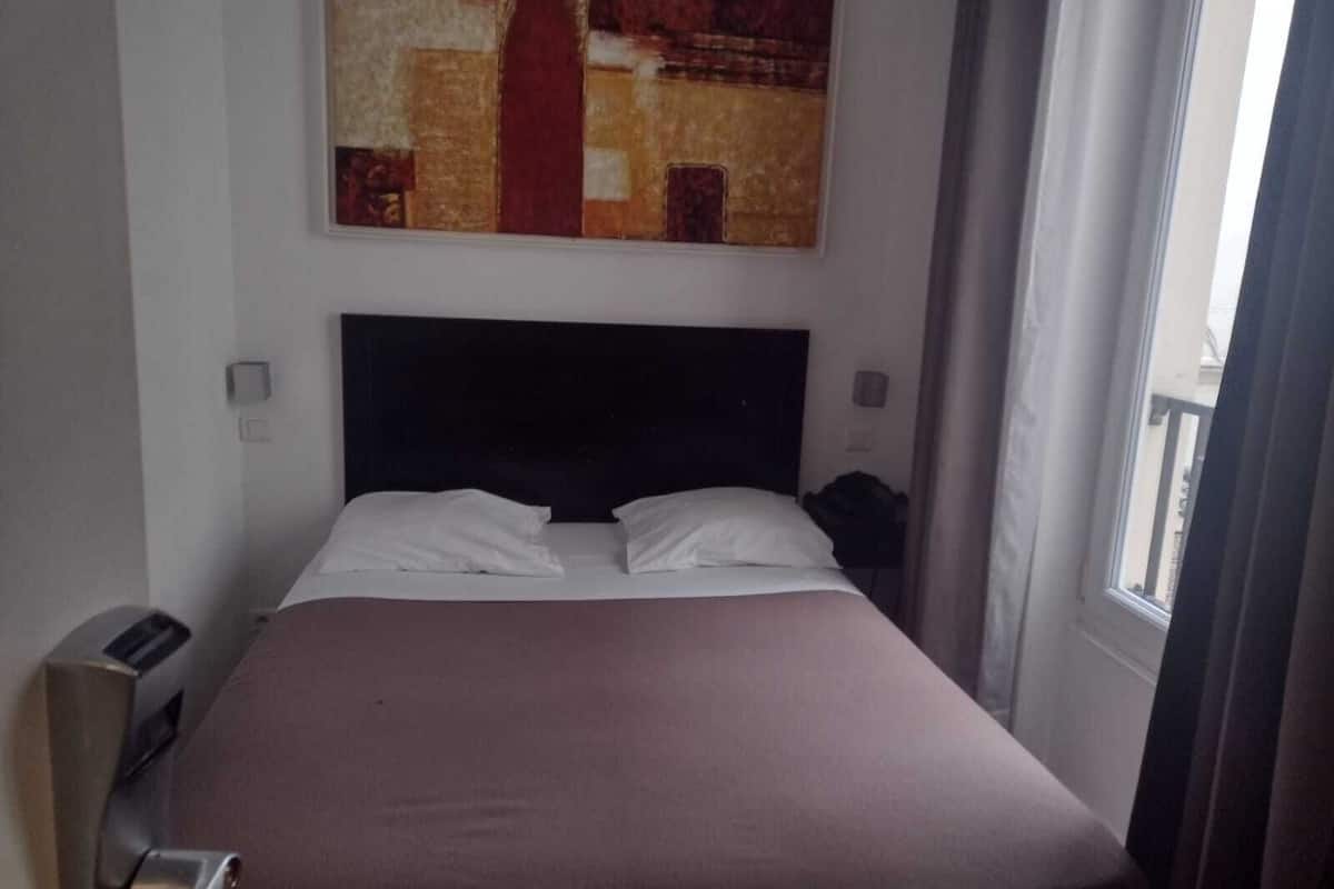 Quarto de hotel com cama, cabeceira, um quadro na parede e janela com cortinas.
