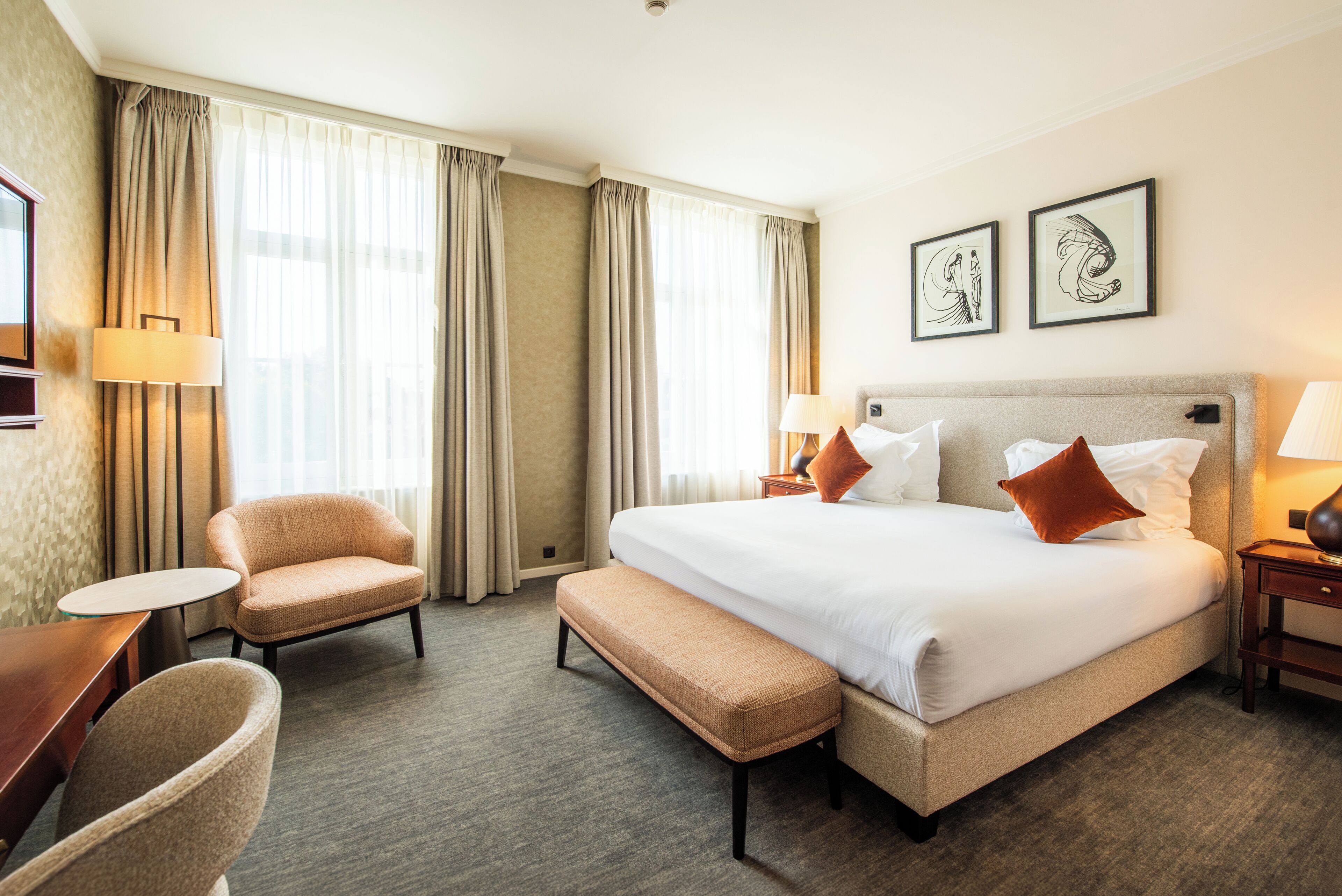 Quarto de hotel com uma cama grande, uma cadeira, um banco, uma escrivaninha e uma televisão.