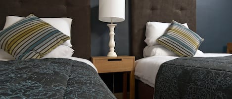 Quarto de hotel com duas camas, cada uma com cabeceira, mesa de cabeceira e almofadas listradas.