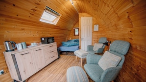 Interior acolhedor de cabana com teto de madeira, cama com roupa de cama estampada, área de estar com poltrona e apoio para os pés, e uma cozinha compacta com cafeteira e micro-ondas.