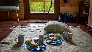 Interior rústico de uma cabana com área de jantar, mesa posta com comida e bebidas, janela com vista para vegetação e paredes de madeira.