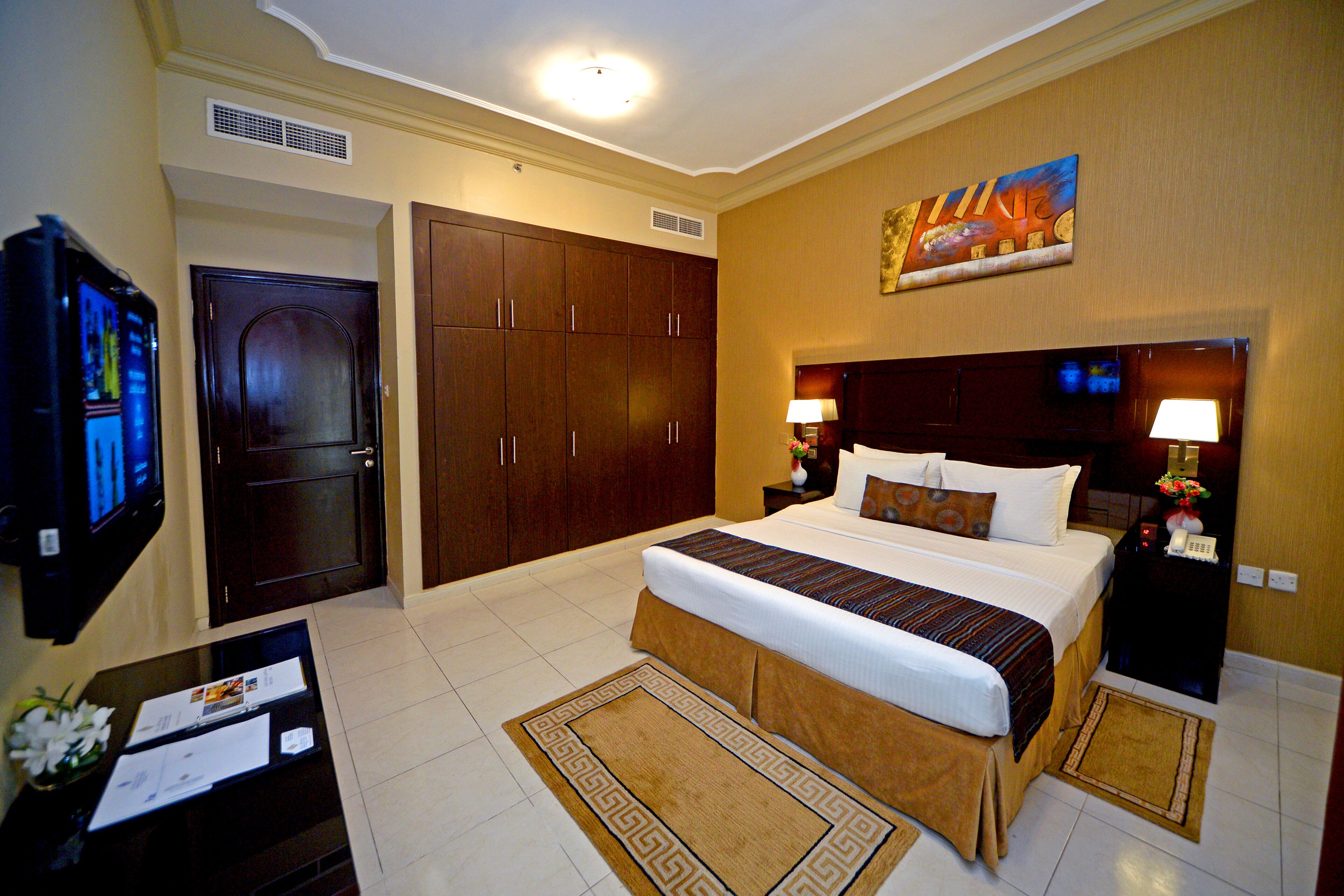 Quarto de hotel com uma cama grande, duas mesas de cabeceira com luminárias, uma TV de tela plana, um guarda-roupa de madeira e um quadro na parede.