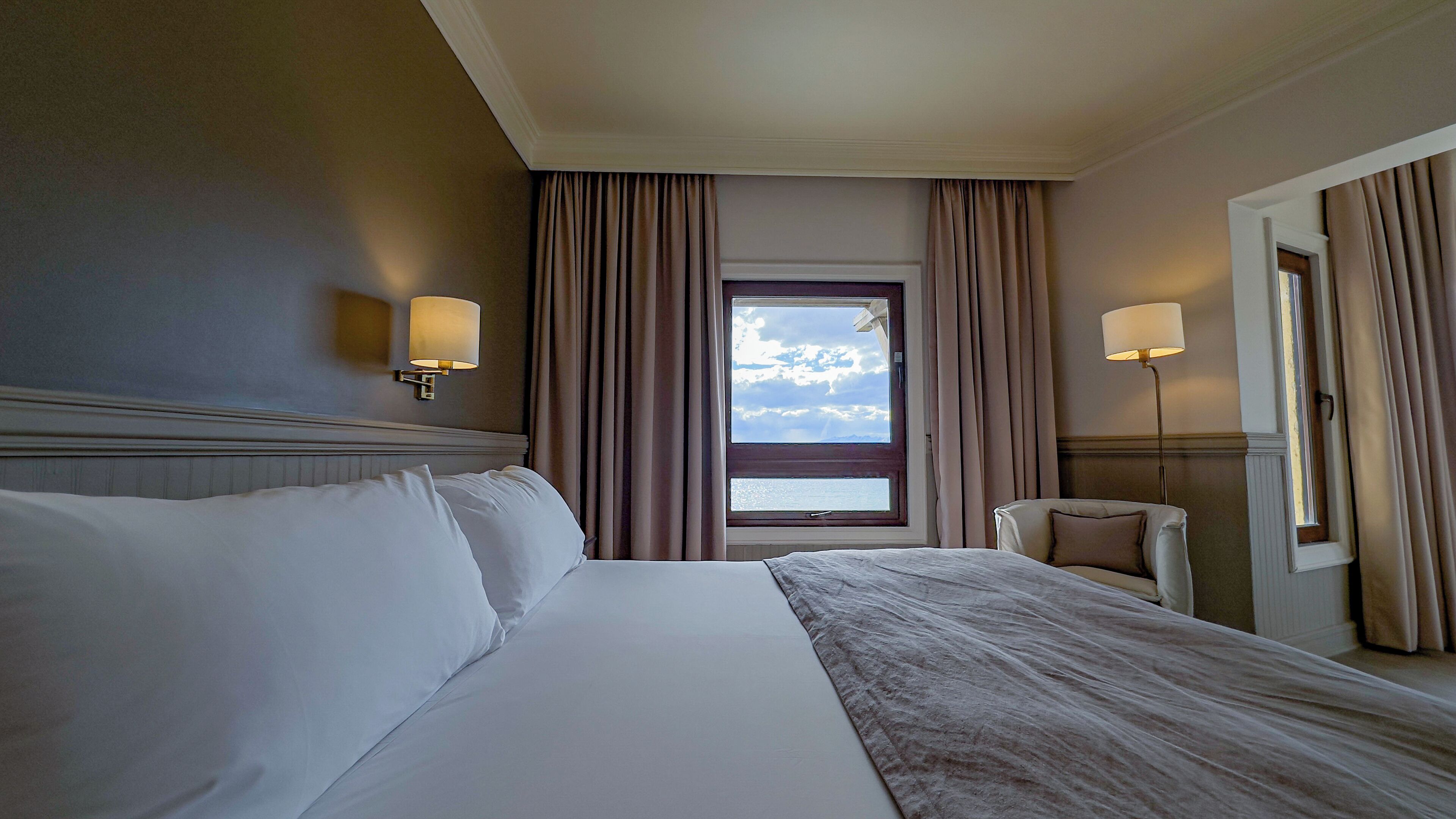 Quarto de hotel com uma cama grande, duas luminárias na parede, uma janela com cortinas e uma cadeira.
