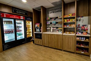 Parte de uma loja com balcões refrigerados identificados como 'Candlewood Coffee Cupboard' e prateleiras repletas de lanches e bebidas.
