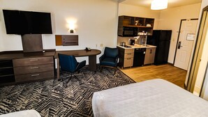 Quarto de hotel com cama, escrivaninha, cadeira, televisão, cozinha compacta e tapete estampado.
