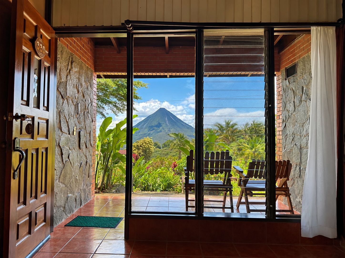 Quarto com porta de vidro deslizante que dá para uma varanda com cadeiras e vista de um vulcão.